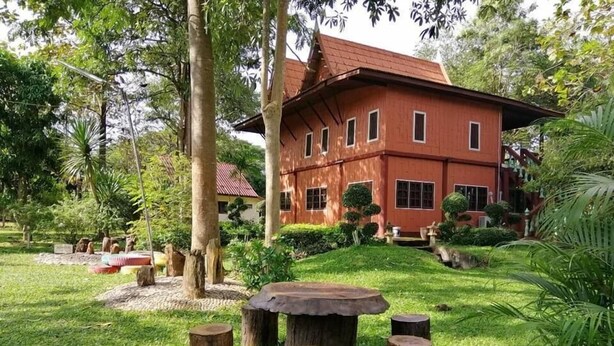 Uma casa vermelha de dois andares com um jardim bem cuidado, incluindo uma mesa e cadeiras de madeira, cercada por árvores e vegetação.