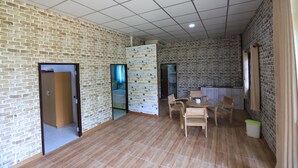 Um quarto com parede de tijolos, teto azulejado, chão de madeira, uma mesa pequena com cadeiras e uma porta que dá para outro cômodo.