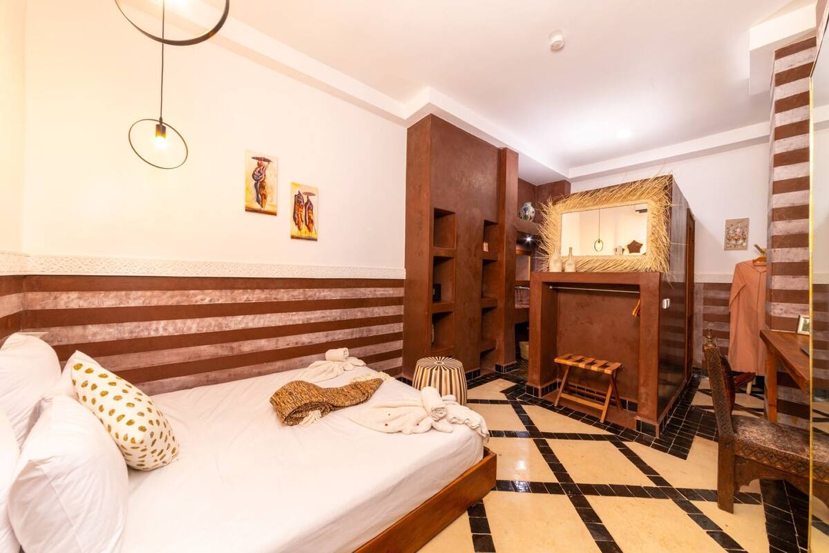 Quarto de hotel com cama, cabeceira de madeira, espelho, um banquinho pequeno e um banco.