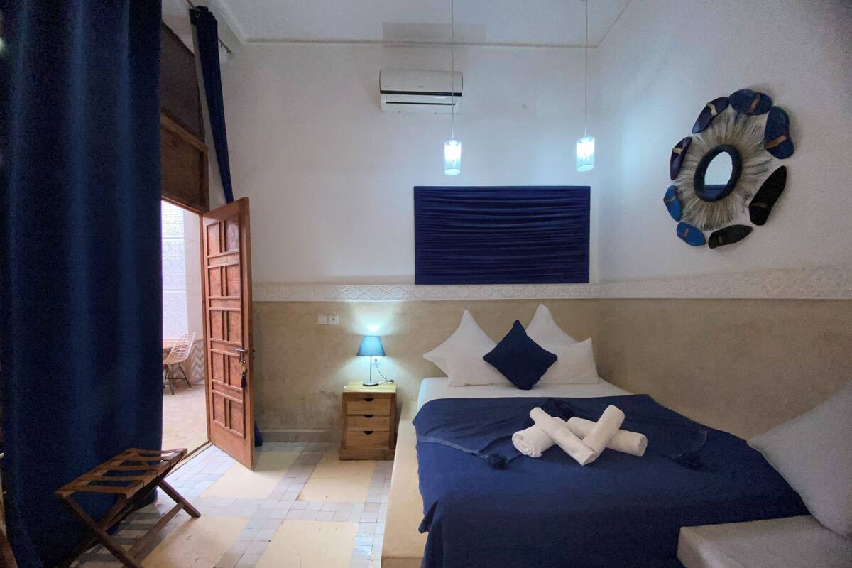Quarto de hotel com uma cama, mesa de cabeceira com abajur, ar-condicionado fixado na parede e um espelho decorativo.