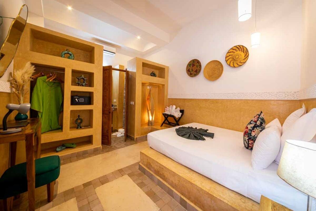 Quarto de hotel com cama, escrivaninha com luminária, cadeira, prateleira com objetos decorativos e porta para outro cômodo.