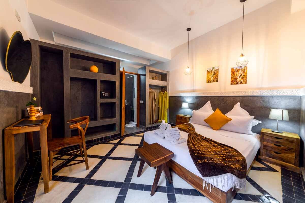 Quarto de hotel com cama, mesas de cabeceira, escrivaninha e cadeira. Guarda-roupa e banheiro visíveis ao fundo.