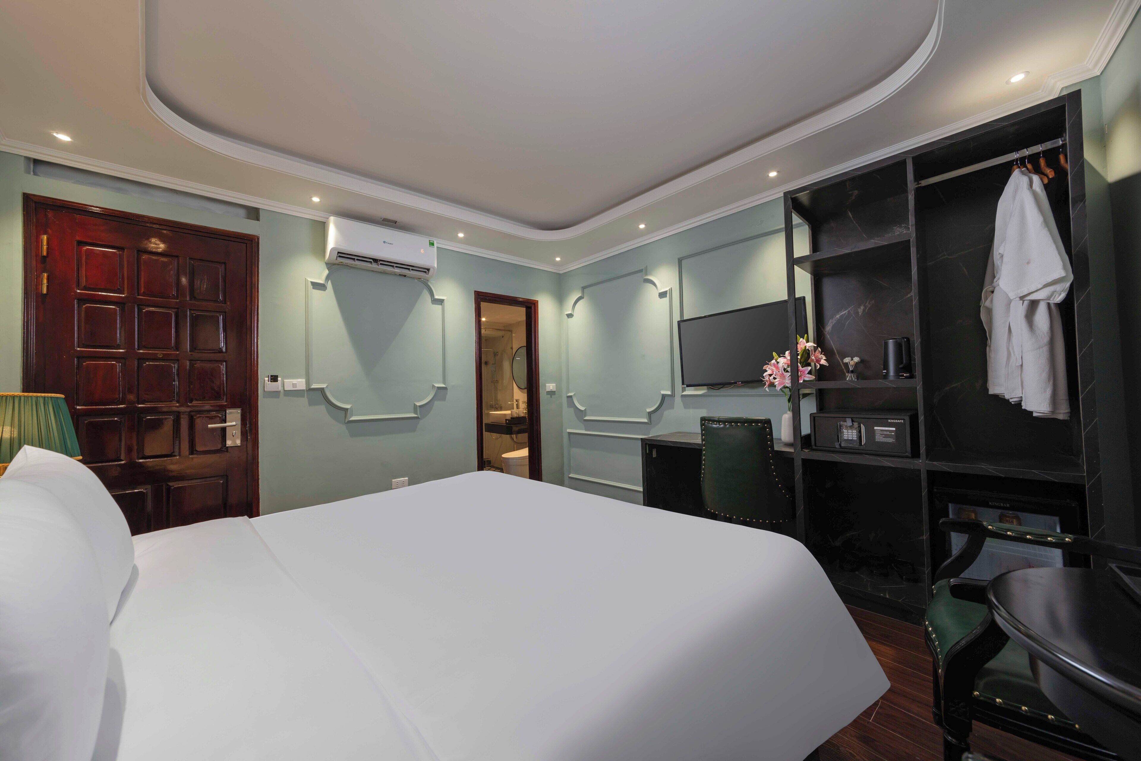 Quarto de hotel com uma cama grande, uma escrivaninha, uma cadeira, uma televisão, um guarda-roupa e um aparelho de ar condicionado.