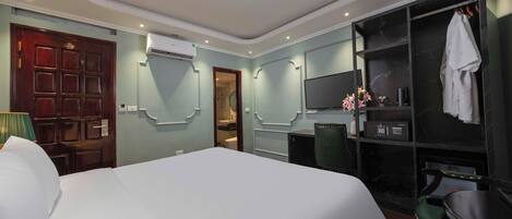 Quarto de hotel com uma cama grande, uma escrivaninha, uma cadeira, uma televisão, um guarda-roupa e um aparelho de ar condicionado.