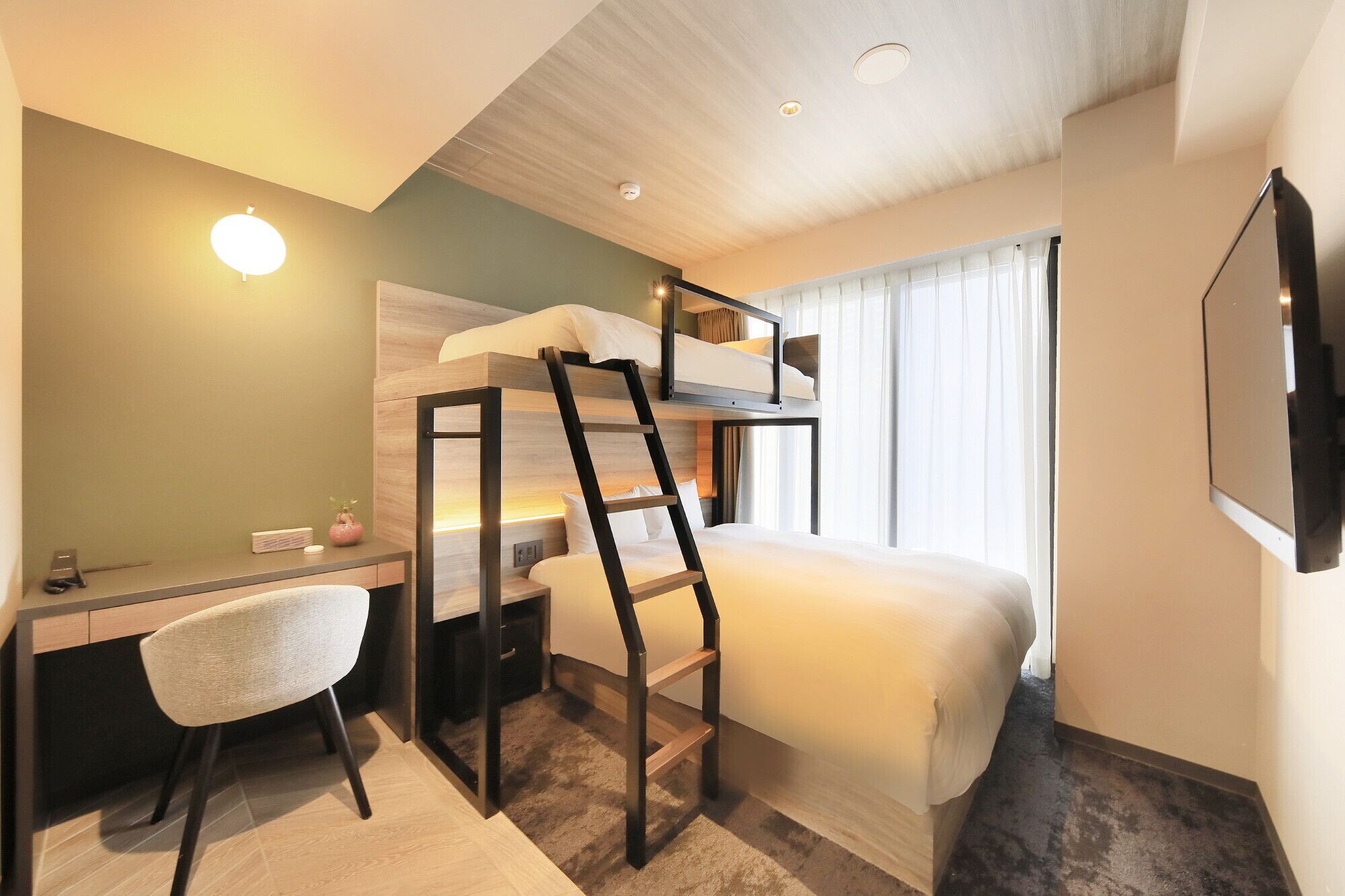 Quarto de hotel com beliches, escrivaninha, cadeira, televisão e janela com cortinas.
