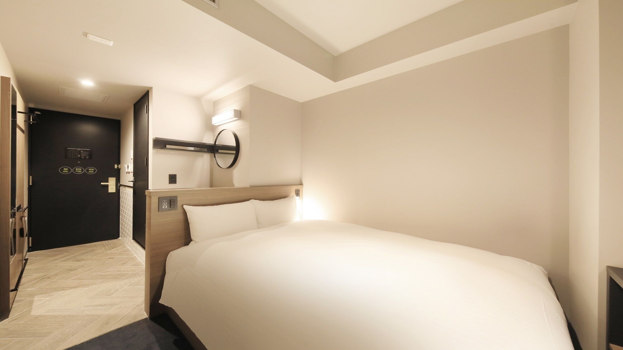 Quarto de hotel com uma cama grande, uma mesa de cabeceira, um espelho fixado na parede e uma porta com painel de controle eletrônico.