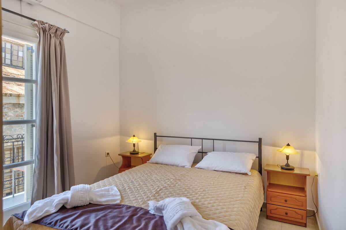 Cama bem arrumada com colcha, duas mesas de cabeceira com abajures, janela com cortinas e vista para prédios lá fora.