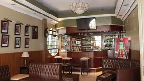 Uma área de bar com balcão, banquetas e uma tela de televisão. Há quadros nas paredes e um lustre sobre o balcão.