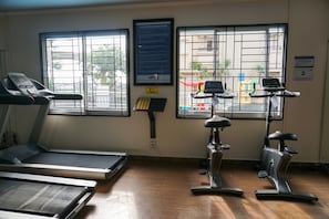 Sala de ginástica com esteiras e bicicletas ergométricas, janelas amplas com barras de metal e um cartaz na parede.