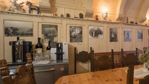 Interior de madeira com uma mesa de jantar, cadeiras, cafeteira, garrafas de vinho e fotografias antigas na parede.