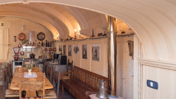 Um interior acolhedor em madeira, com uma longa mesa de jantar, bancos e uma lareira central.
