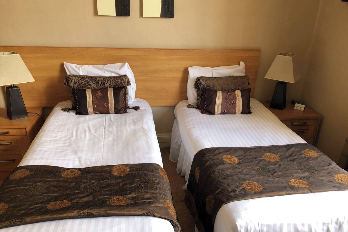 Duas camas de solteiro com cabeceiras, roupa de cama branca e almofadas decorativas. Cada cama possui uma mesa de cabeceira com abajur.