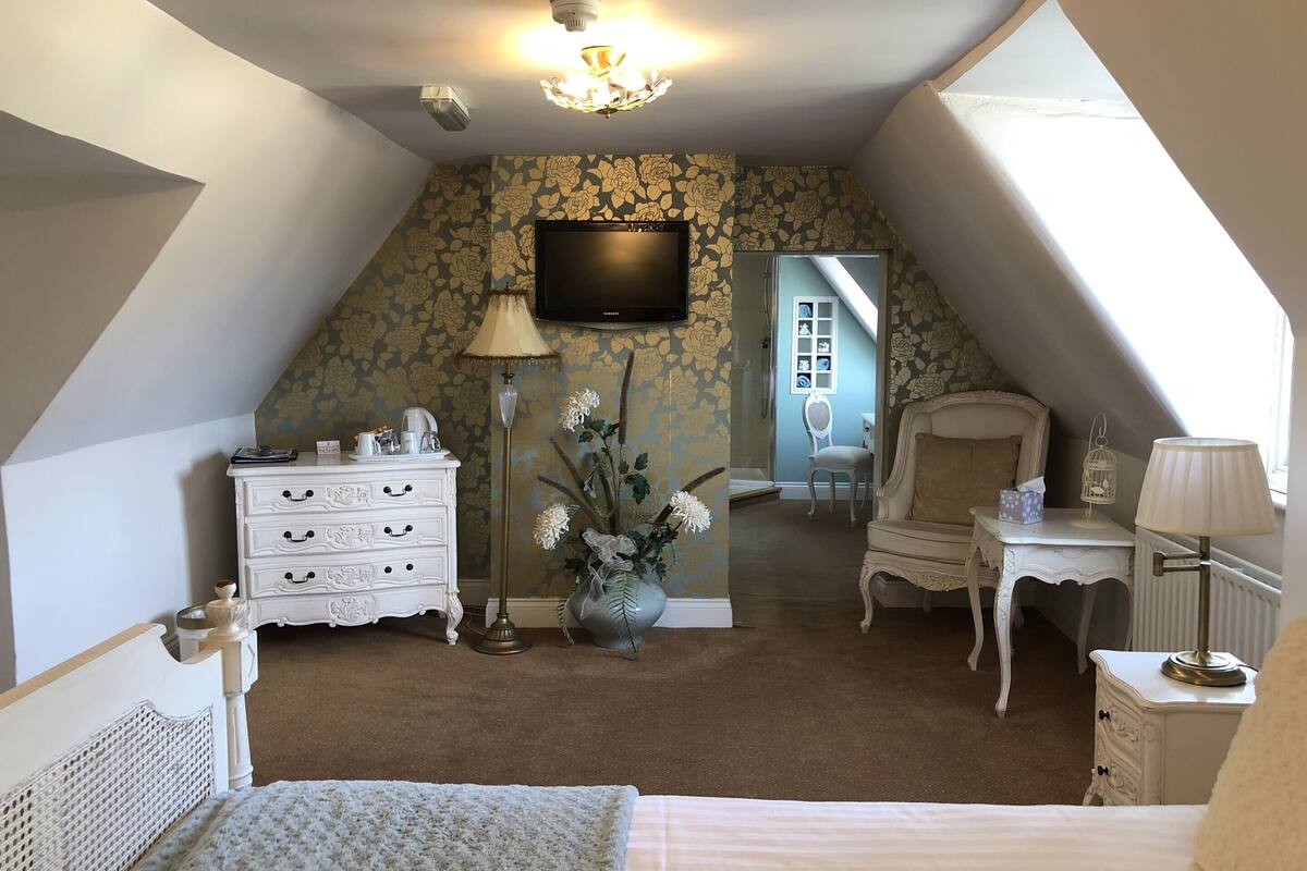 Quarto com teto inclinado, televisão na parede, cómoda, cadeira, mesa de cabeceira e candeeiro.