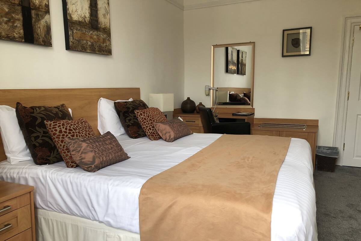 Quarto de hotel com uma cama grande, uma escrivaninha, uma cadeira, um espelho e obras de arte nas paredes.