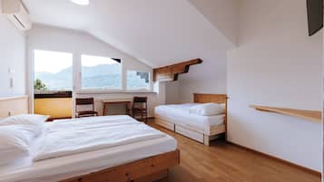 Quarto com duas camas, uma viga de madeira no teto, um radiador e janela com vista para as montanhas.
