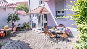 Um pátio com mesa e cadeiras, um pequeno jardim e um edifício com telhado de telhas vermelhas.