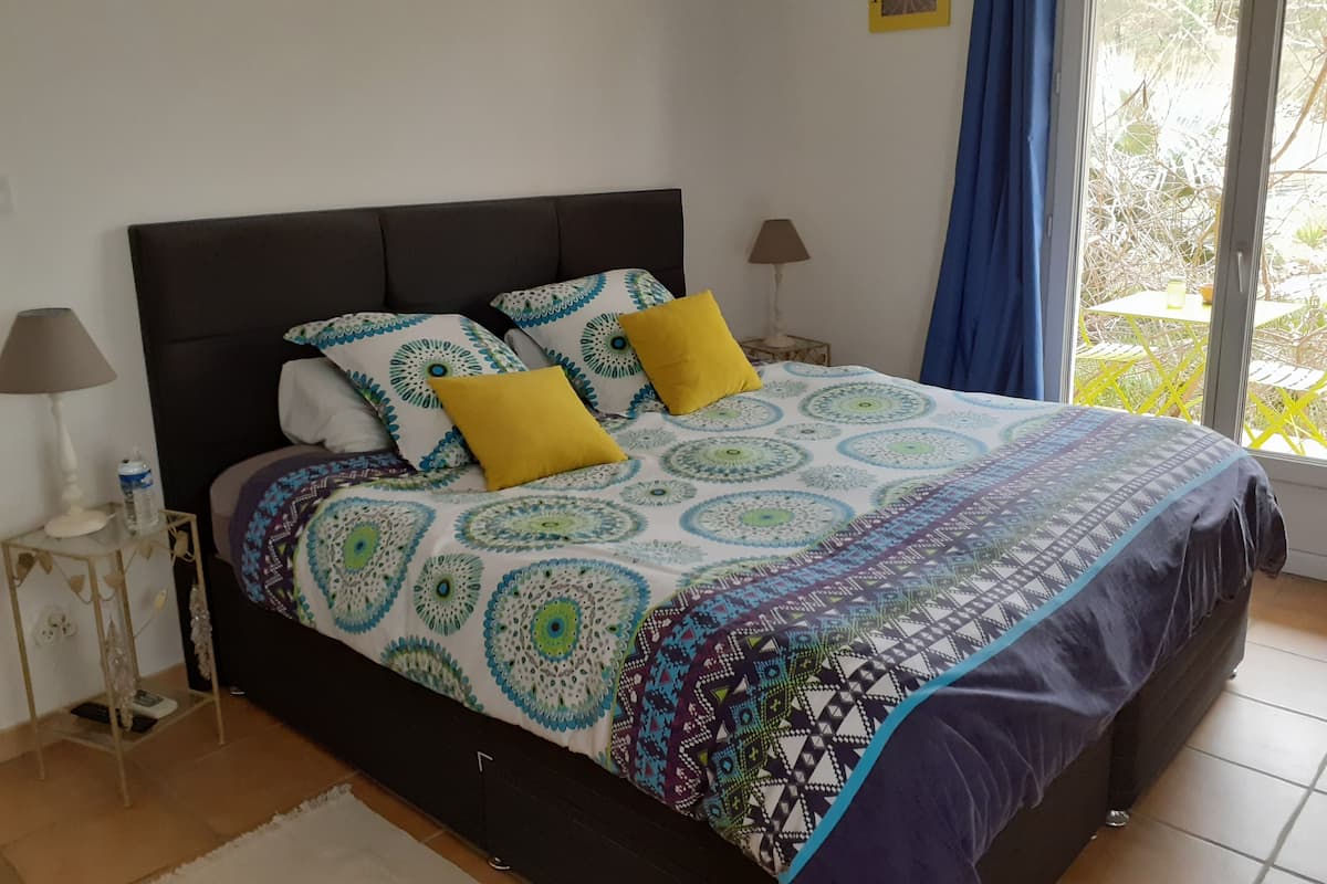 Quarto com cama, almofadas, mesa de cabeceira, luminária e janela com cortinas.