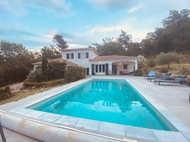 Uma piscina com deck ladrilhado, uma casa com telhado telhado e uma área de varanda com cadeiras de estar.