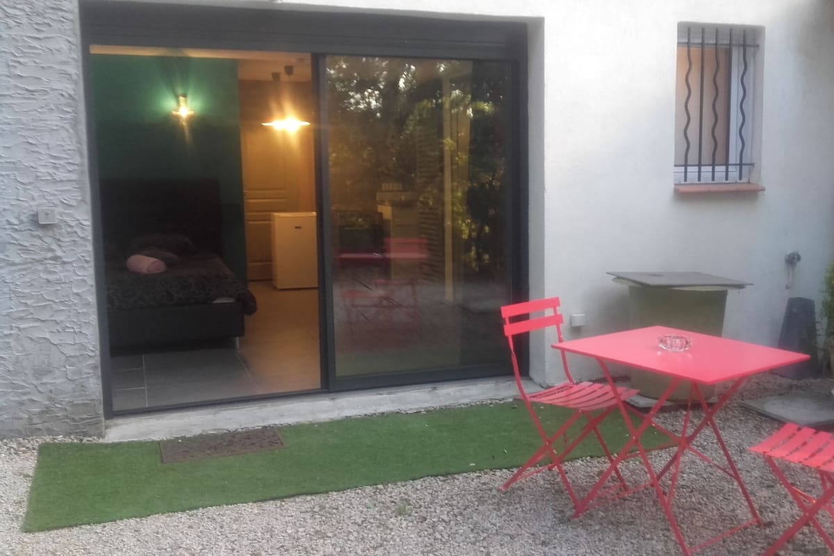 Um pátio com mesa e cadeiras vermelhas, um pequeno jardim e uma porta de correr que dá para um quarto com cama e geladeira.