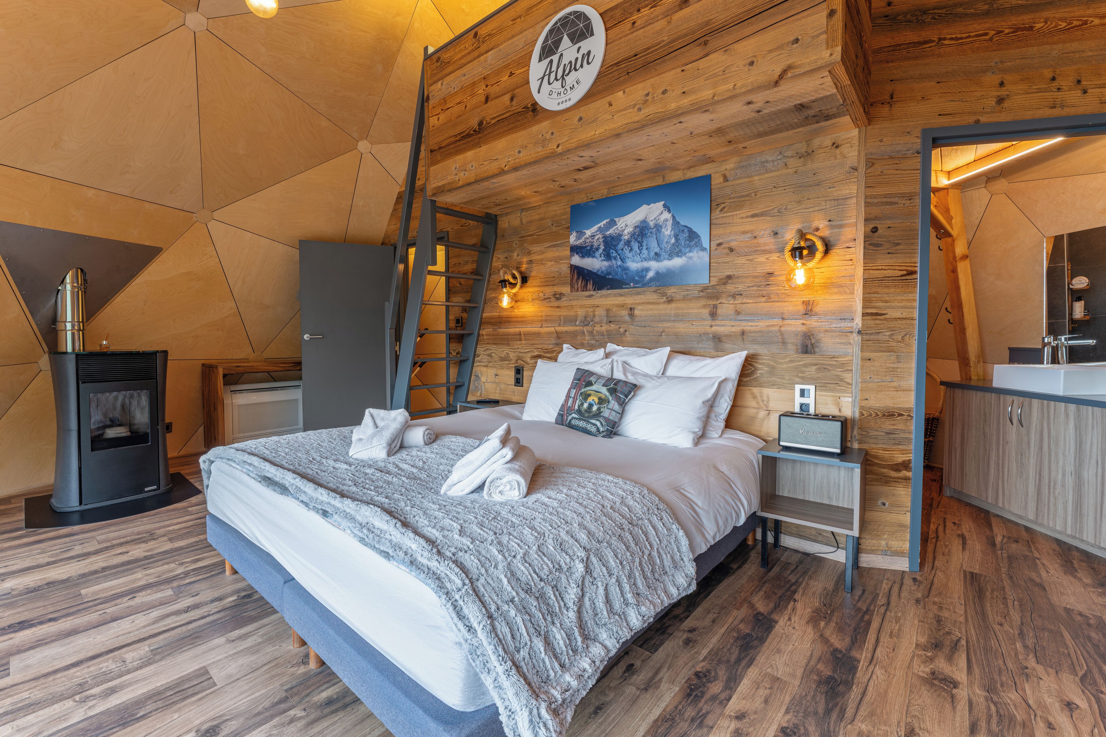 Quarto aconchegante com teto de madeira, uma cama com cobertor, mesa de cabeceira e luminária na parede.