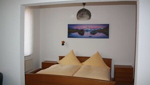 Quarto com cama de madeira, duas almofadas, mesa de cabeceira, luminária na parede e pintura de uma paisagem.