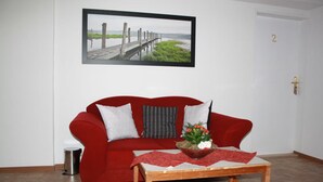 Um sofá vermelho com almofadas brancas e cinzentas, uma mesa de centro de madeira com um jogo americano vermelho e uma moldura com uma fotografia de um cais na parede.
