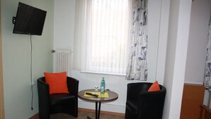 Quarto com duas cadeiras pretas, uma mesa redonda pequena, televisão fixada na parede, janela com cortinas leves e um radiador.