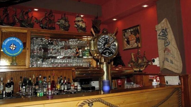Um bar com um relógio, prateleiras cheias de garrafas e uma parede vermelha decorada com motivos náuticos.