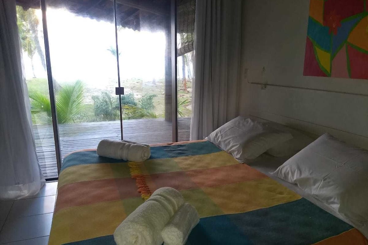 Uma cama com roupa de cama colorida, uma janela com cortinas e vista para o verde lá fora.