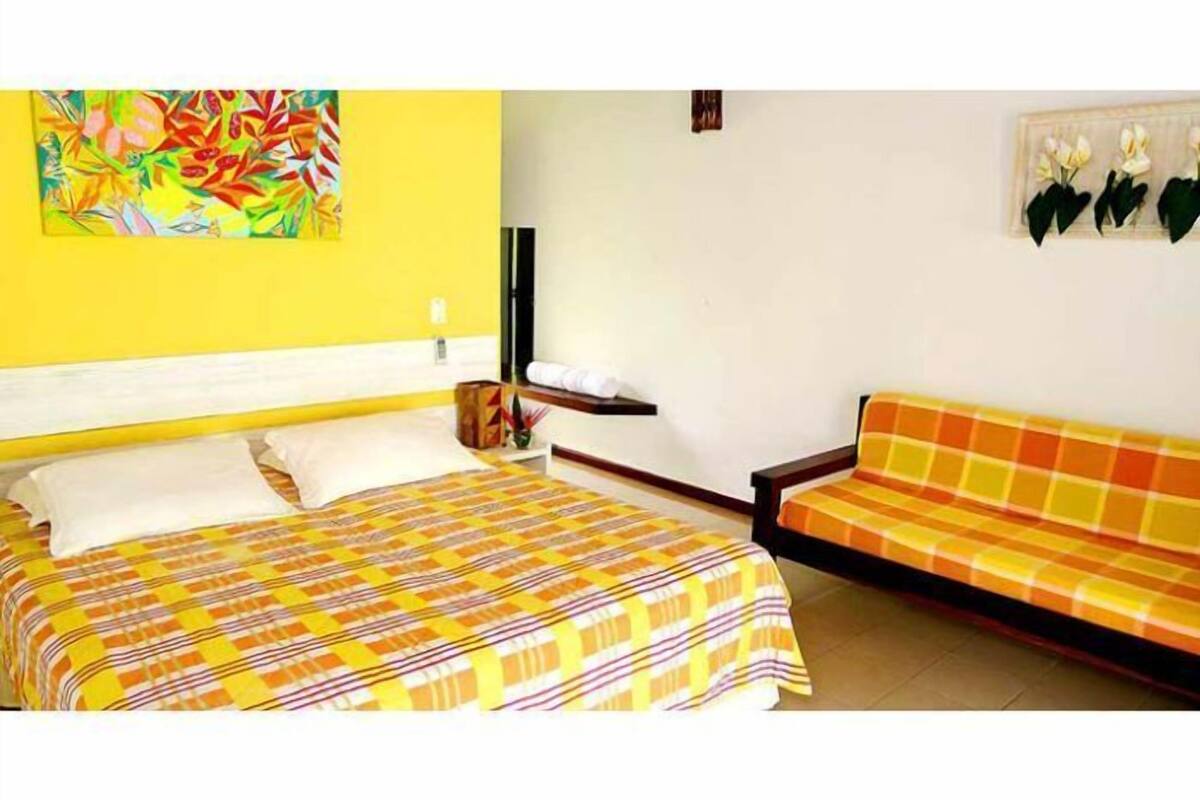 Quarto com cama, sofá, quadro na parede e televisão fixada na parede.