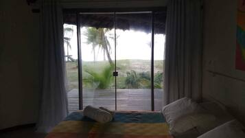 Quarto com cama, vista para palmeiras e uma praia através de grandes janelas, e um deck de madeira na área externa.