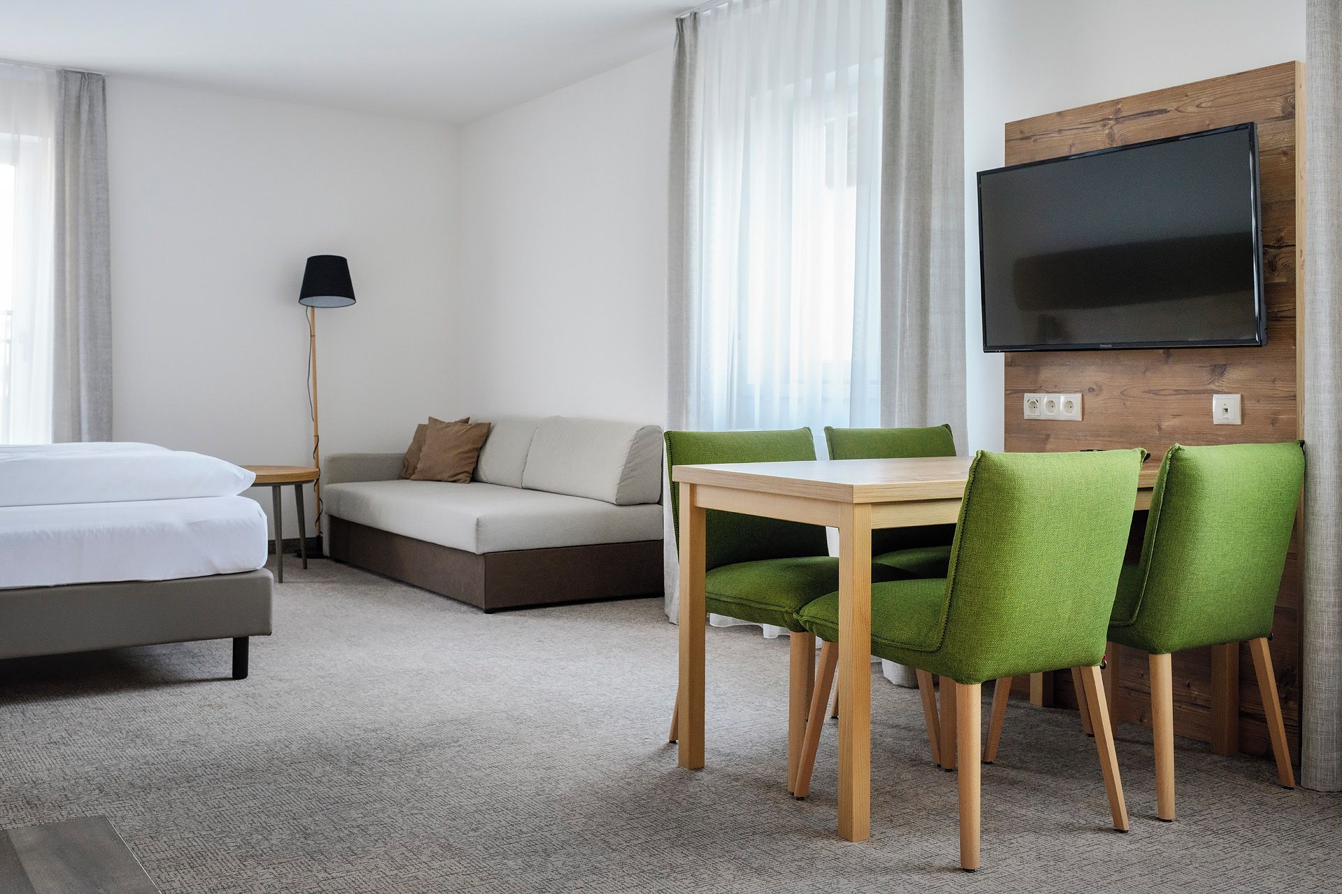 Quarto de hotel com cama, sofá, mesa de jantar com cadeiras, televisão presa à parede e abajur.