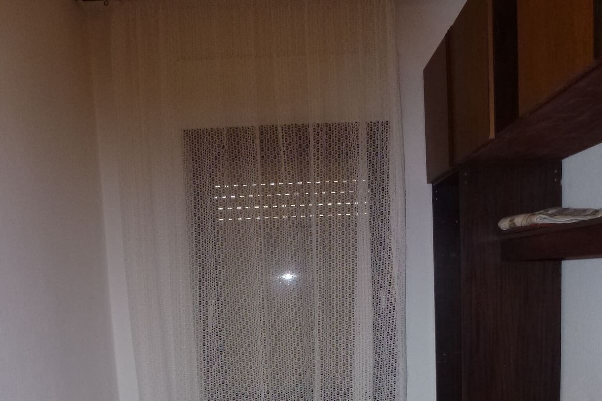 Quarto pequeno com uma cama de solteiro, cabeceira de madeira, janela com cortina de renda e radiador.