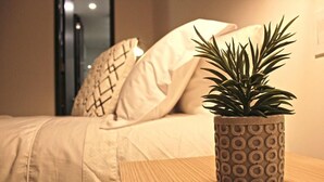 Uma cama com edredom bege e almofadas estampadas, uma pequena planta em vaso sobre uma mesa de cabeceira de madeira, e um espelho refletindo o quarto.