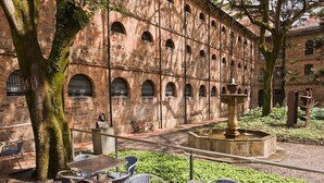 Um pátio externo com mesas e cadeiras, uma fonte e um edifício de tijolos com janelas em arco.