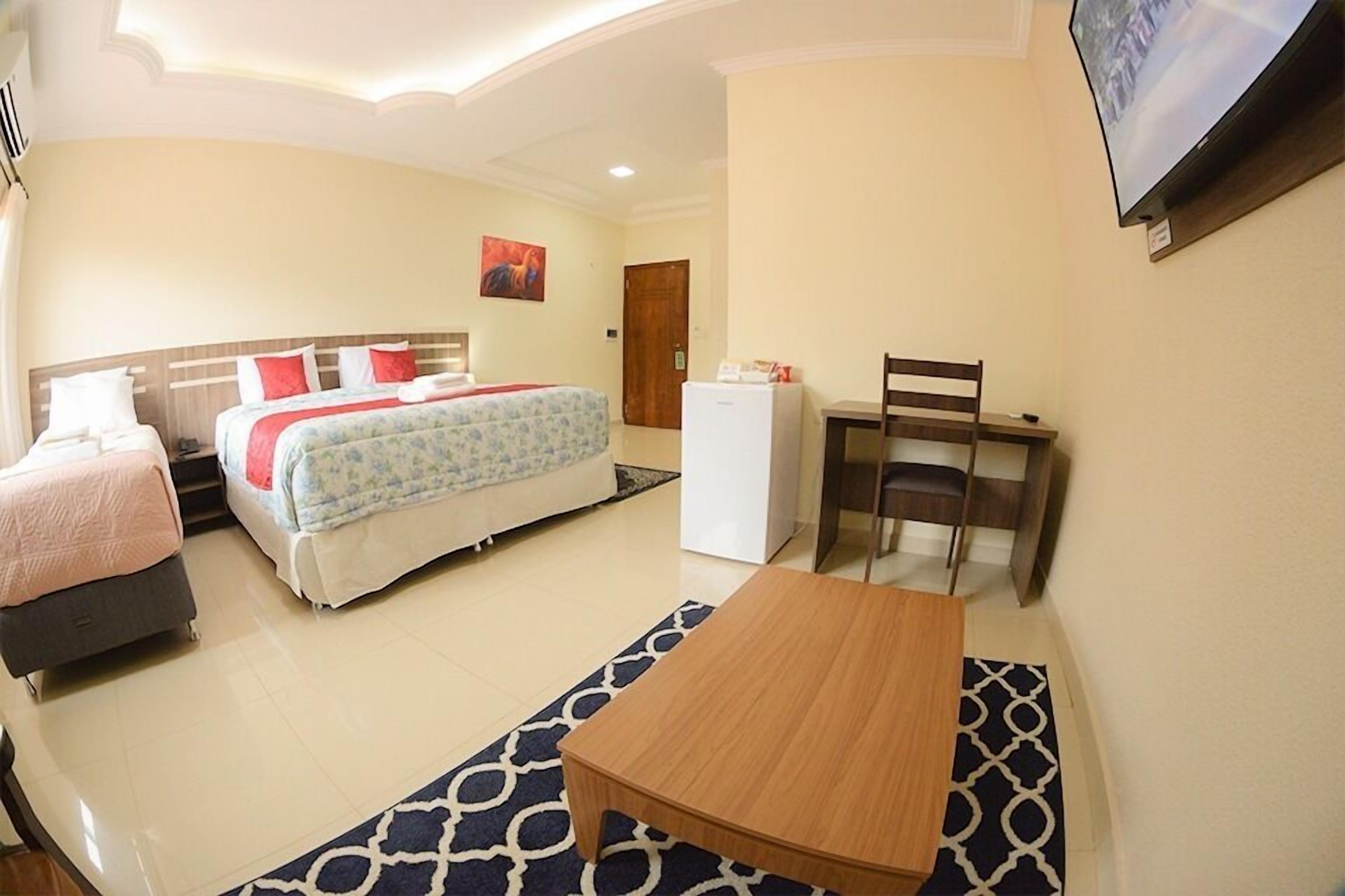 Quarto de hotel com duas camas, uma mesa, uma cadeira, uma televisão e um frigorífico.