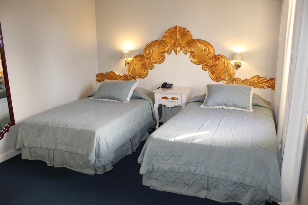 Quarto de hotel com duas camas, cada uma com colcha e almofadas azul-claras, uma mesinha de cabeceira branca com telefone e uma cabeceira dourada decorativa.