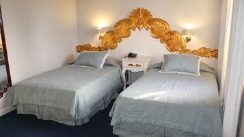 Quarto de hotel com duas camas, cada uma com colcha e almofadas azul-claras, uma mesinha de cabeceira branca com telefone e uma cabeceira dourada decorativa.