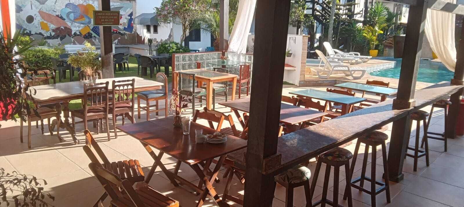 Uma área de refeições ao ar livre com mesas e cadeiras de madeira, uma piscina e um mural na parede.