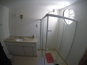 Uma casa de banho com cabine de duche em vidro, lavatório com bancada e uma pequena janela.