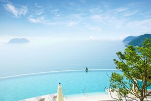Vista nítida de uma piscina infinita com alguém nadando, rodeado por espreguiçadeiras e uma árvore.