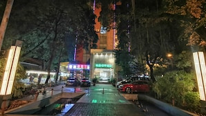 Vista noturna de um edifício com sinalização iluminada, área de entrada pavimentada e vegetação ao redor.