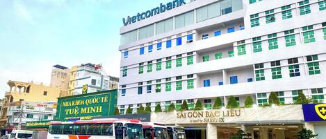Um edifício de vários andares com uma placa chamativa da 'Wetcombank', um hotel chamado 'Sài Gòn Bac Lieu', e vários ônibus e carros estacionados na frente.