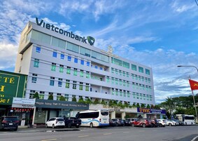 Um edifício de vários andares com um destaque para o letreiro 'Vietcombank', uma bandeira e uma placa indicando 'Ngân hàng TMCP Nghiệp thượng Việt Nam'.
