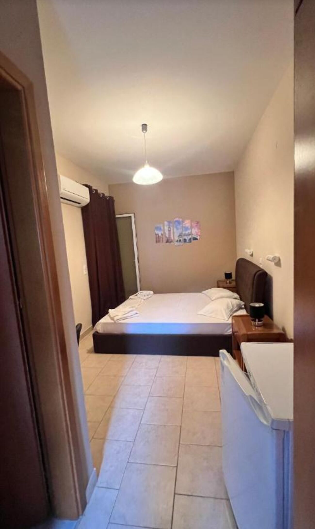 Quarto pequeno e limpo, com cama, mesa de cabeceira, ar-condicionado fixado na parede e luz no teto.