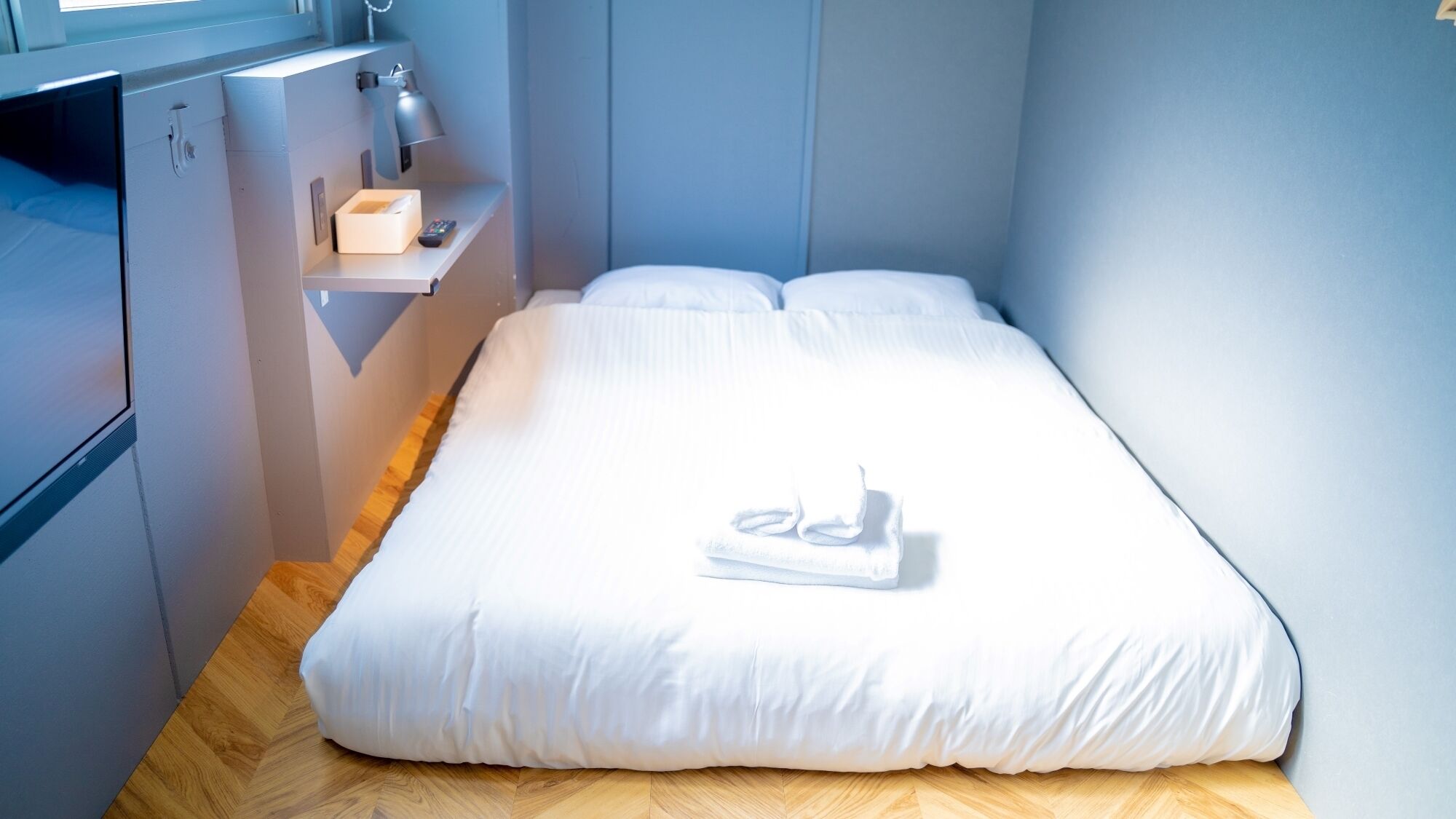 Uma cama pequena e bem arrumada com lençóis brancos e uma toalha dobrada, uma mesa de cabeceira com abajur e controle remoto, e um piso de madeira.
