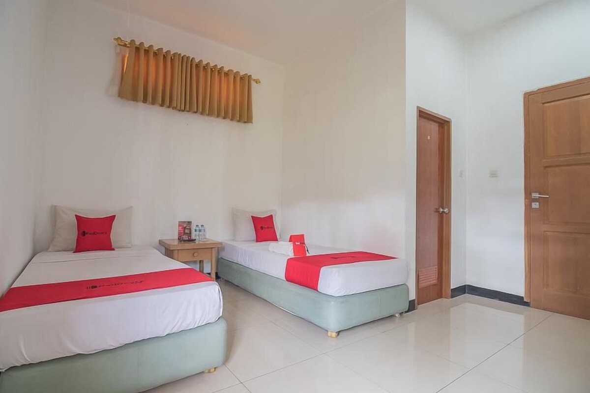 Quarto de hotel com duas camas, uma mesinha de cabeceira, uma porta de madeira e uma janela com cortinas.