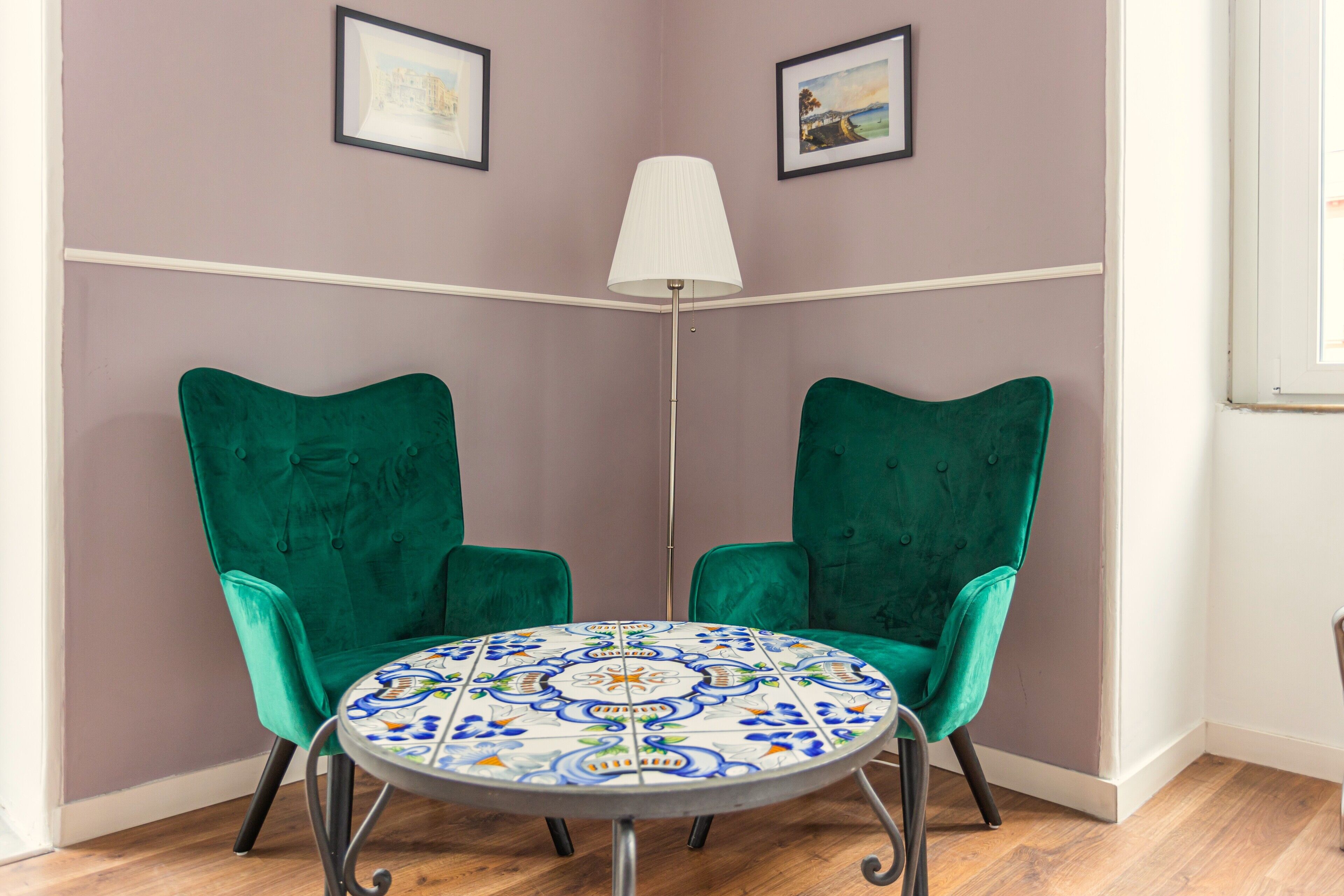 Quarto com uma mesa redonda, duas poltronas estofadas em verde, um abajur e duas imagens emolduradas na parede.