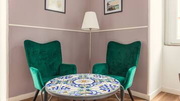 Quarto com uma mesa redonda, duas poltronas estofadas em verde, um abajur e duas imagens emolduradas na parede.