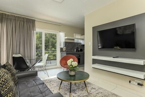 Uma sala de estar moderna com um sofá, uma mesa de jantar com cadeiras, uma TV fixada na parede e uma cozinha visível por uma porta aberta.